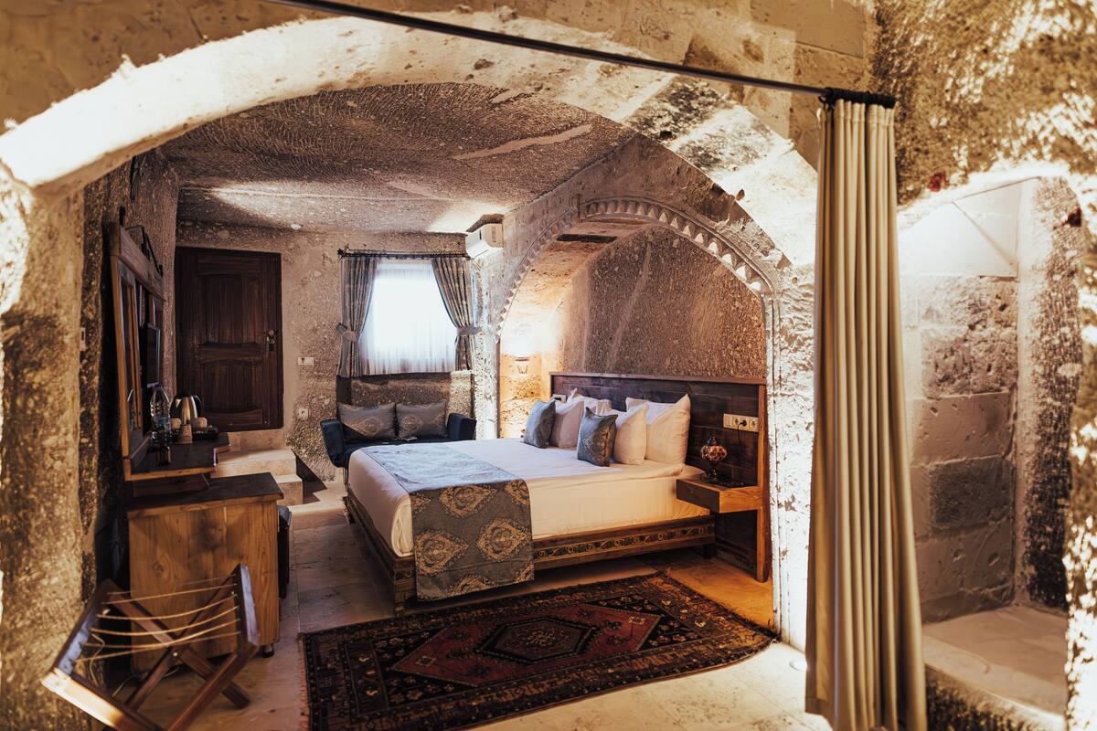 Um quarto rústico com um arco de pedra, uma cama de madeira com roupa de cama estampada, uma mesa de cabeceira com abajur e uma pequena escrivaninha de madeira.