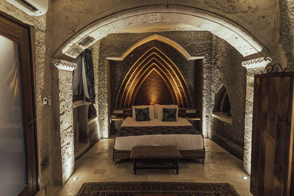 Quarto com arco de pedra, cama com almofadas, um banco e uma mesa de cabeceira.