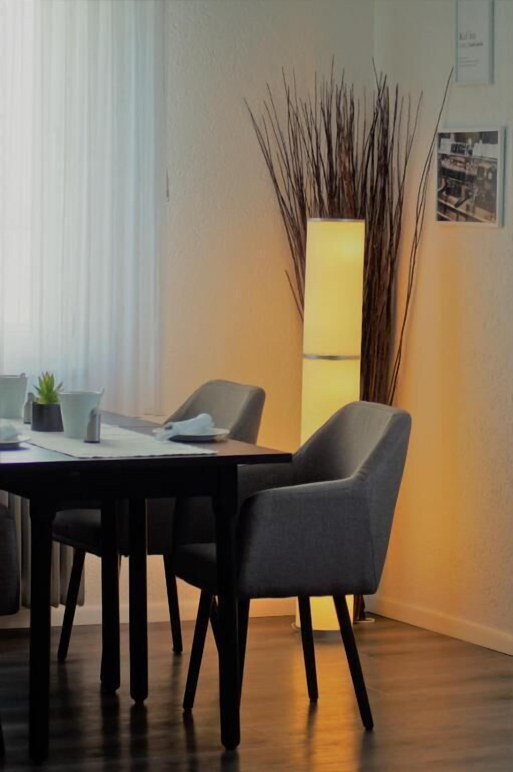 Uma zona de refeições moderna com uma mesa de madeira escura, duas cadeiras estofadas cinzentas, uma pequena planta em vaso e um candeeiro de chão alto com cúpula amarela.