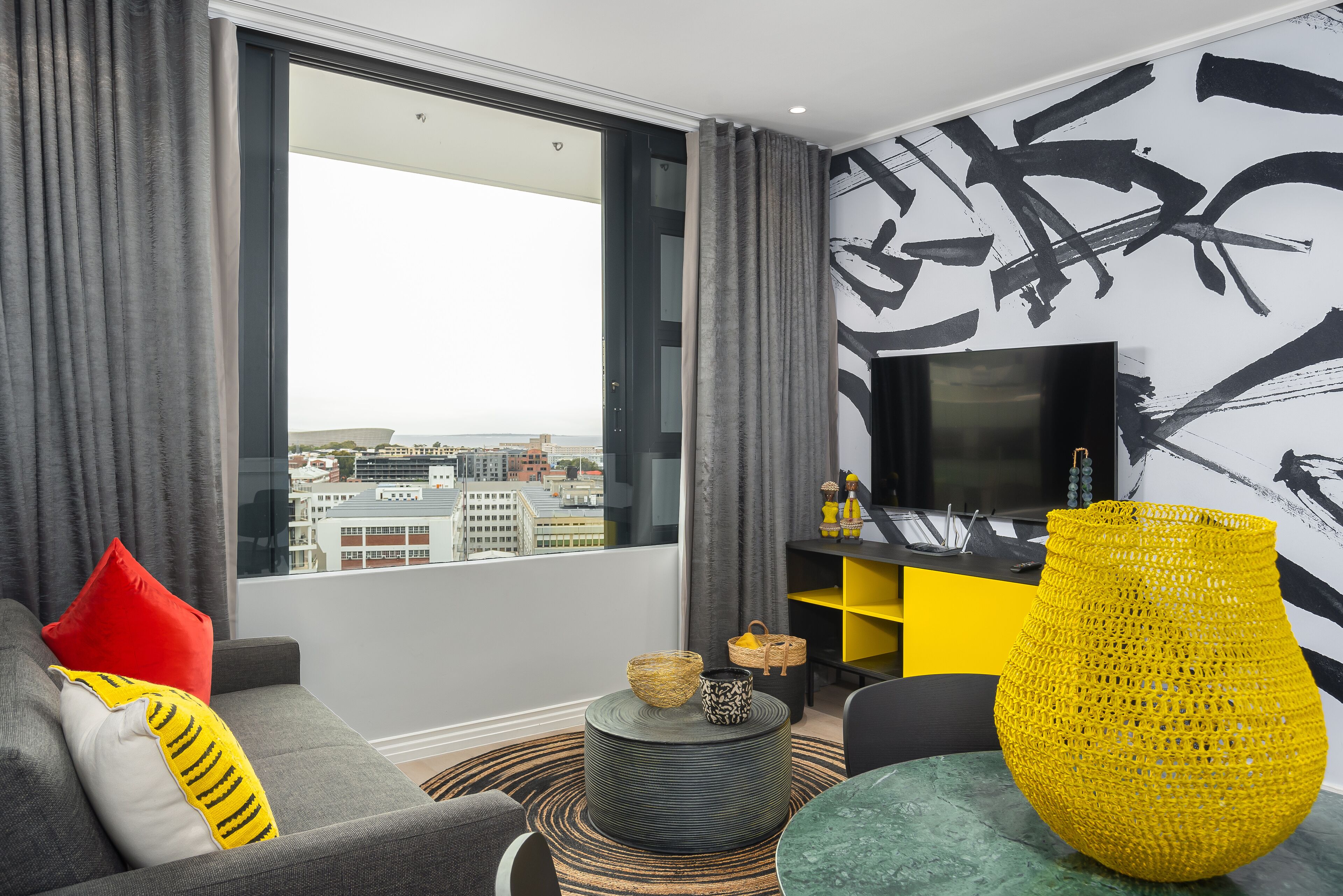 Uma sala de estar moderna com um sofá cinzento, almofadas vermelhas e amarelas, um vaso trançado amarelo e um móvel de entretenimento preto e amarelo.