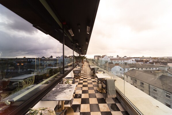 Varanda com piso xadrez, mesas e cadeiras, com vista para um horizonte urbano com edifícios e céu nublado.