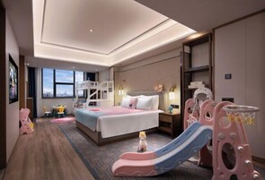 Quarto de hotel com cama, escrivaninha, cadeira, mesinha, estante de livros, janela e espaço infantil.