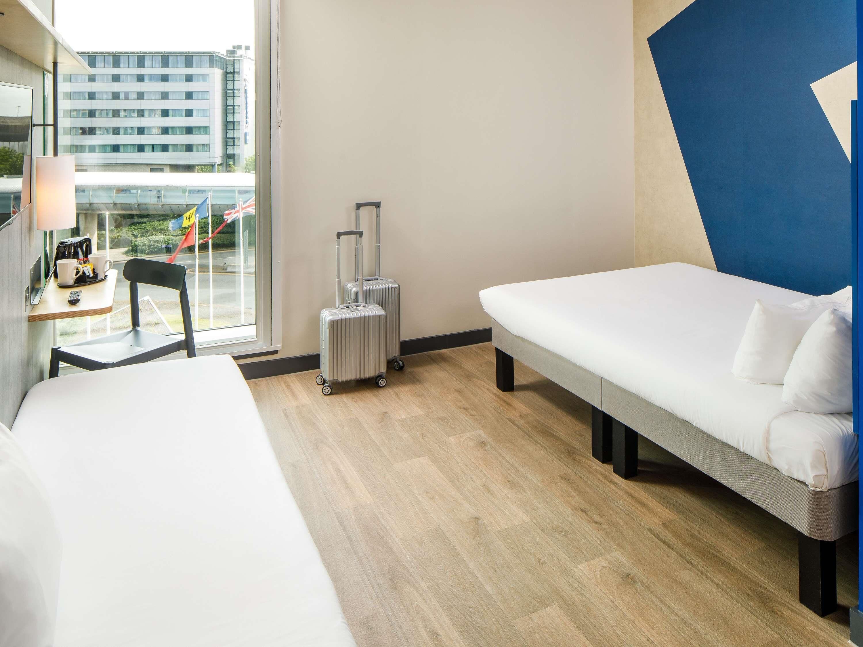 Quarto de hotel com cama, escrivaninha com cadeira, mala e vista para um edifício.