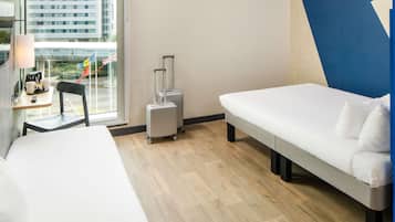 Quarto de hotel com cama, escrivaninha com cadeira, mala e vista para um edifício.