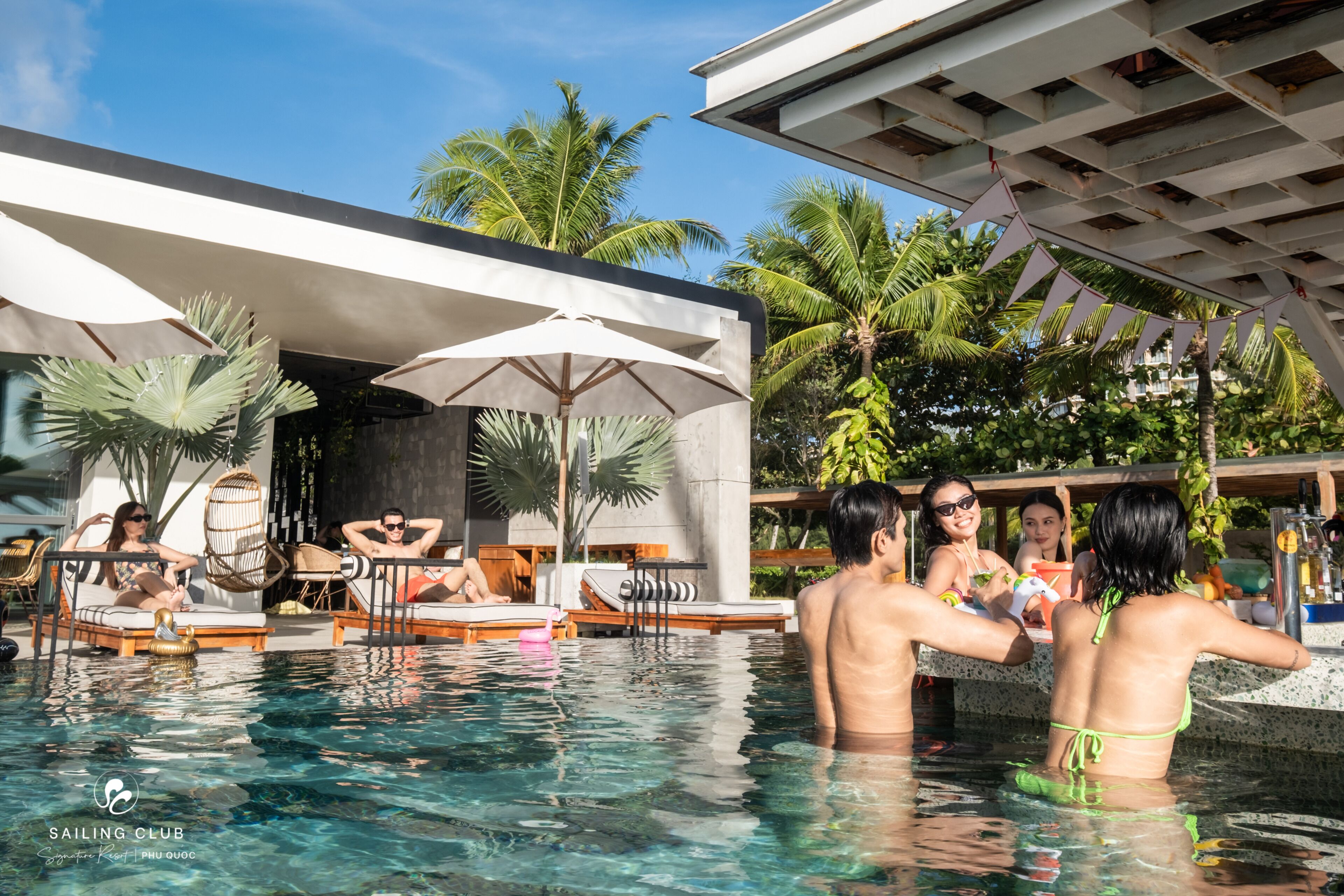 Área de piscina com espreguiçadeiras e guarda-sóis, cercada por palmeiras e uma estrutura de edifício moderna.