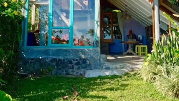 Uma casa rústica com telhado de palha, base de pedra e uma grande janela com detalhes azuis.