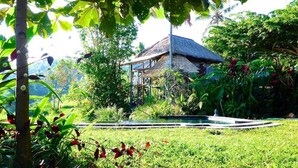 Uma casa rústica com telhado de palha, rodeada por vegetação exuberante e um pequeno lago.