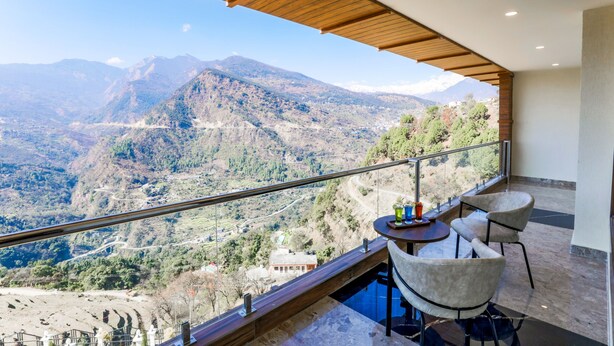 Varanda com vista para uma paisagem montanhosa, mesa com cadeiras e guarda-corpo de vidro.