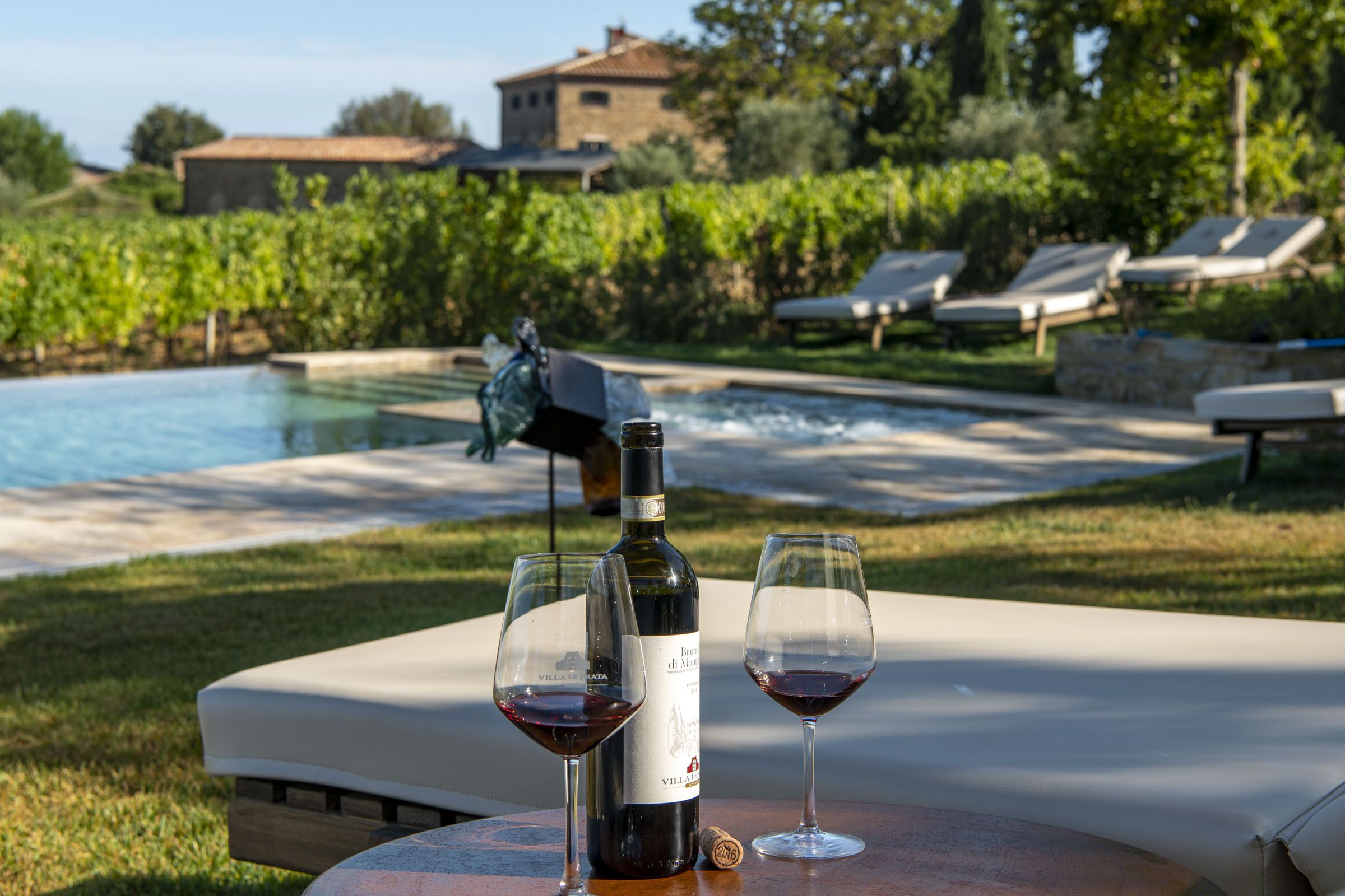 Um ambiente à beira da piscina com uma garrafa de vinho e duas taças sobre uma mesa, cercado por cadeiras de descanso e um vinhedo ao fundo.