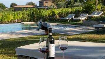 Um ambiente à beira da piscina com uma garrafa de vinho e duas taças sobre uma mesa, cercado por cadeiras de descanso e um vinhedo ao fundo.