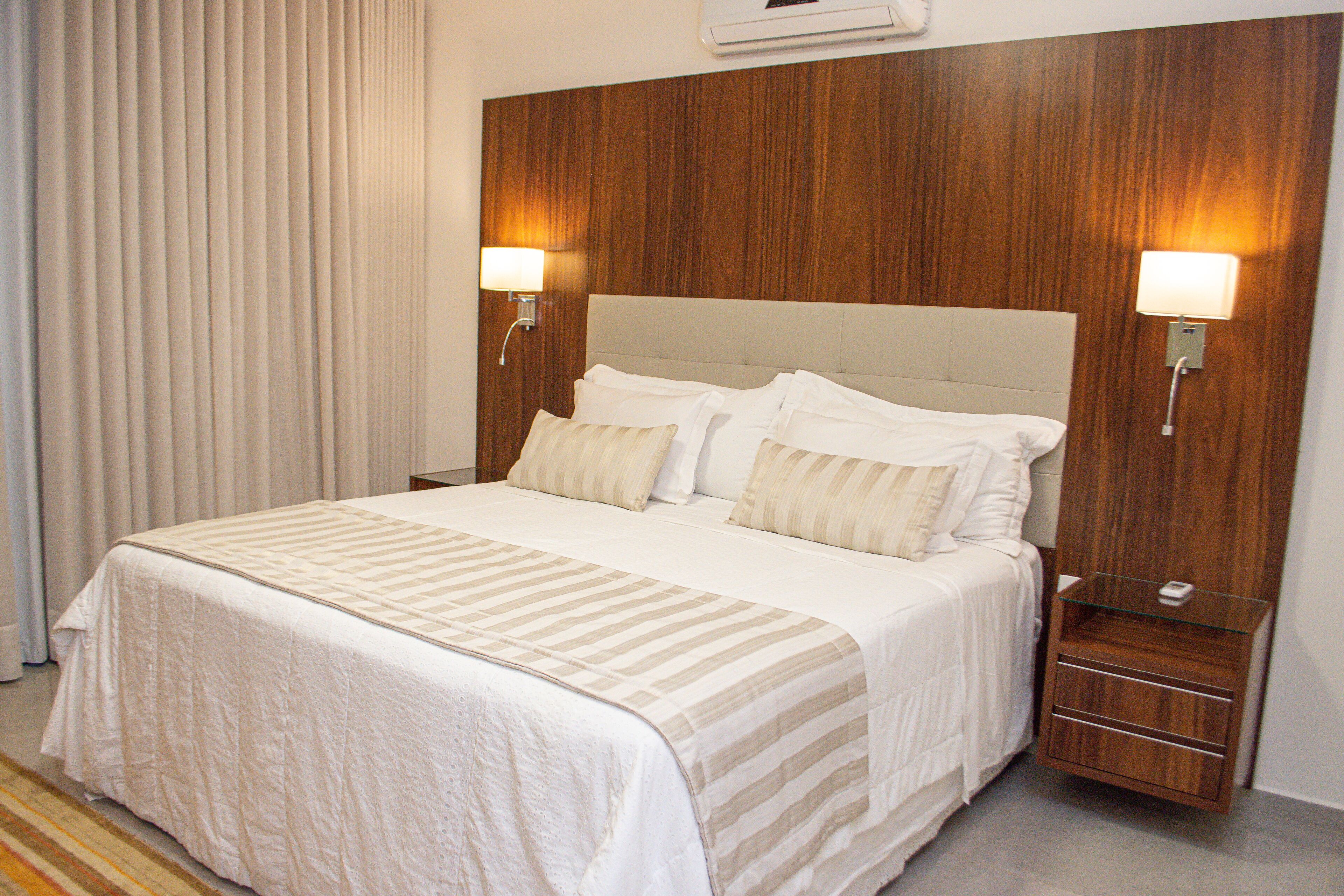 Quarto de hotel com uma cama grande, duas luminárias de cabeceira, um painel de madeira na cabeceira e uma mesa de cabeceira com um telefone e uma gavetinha.