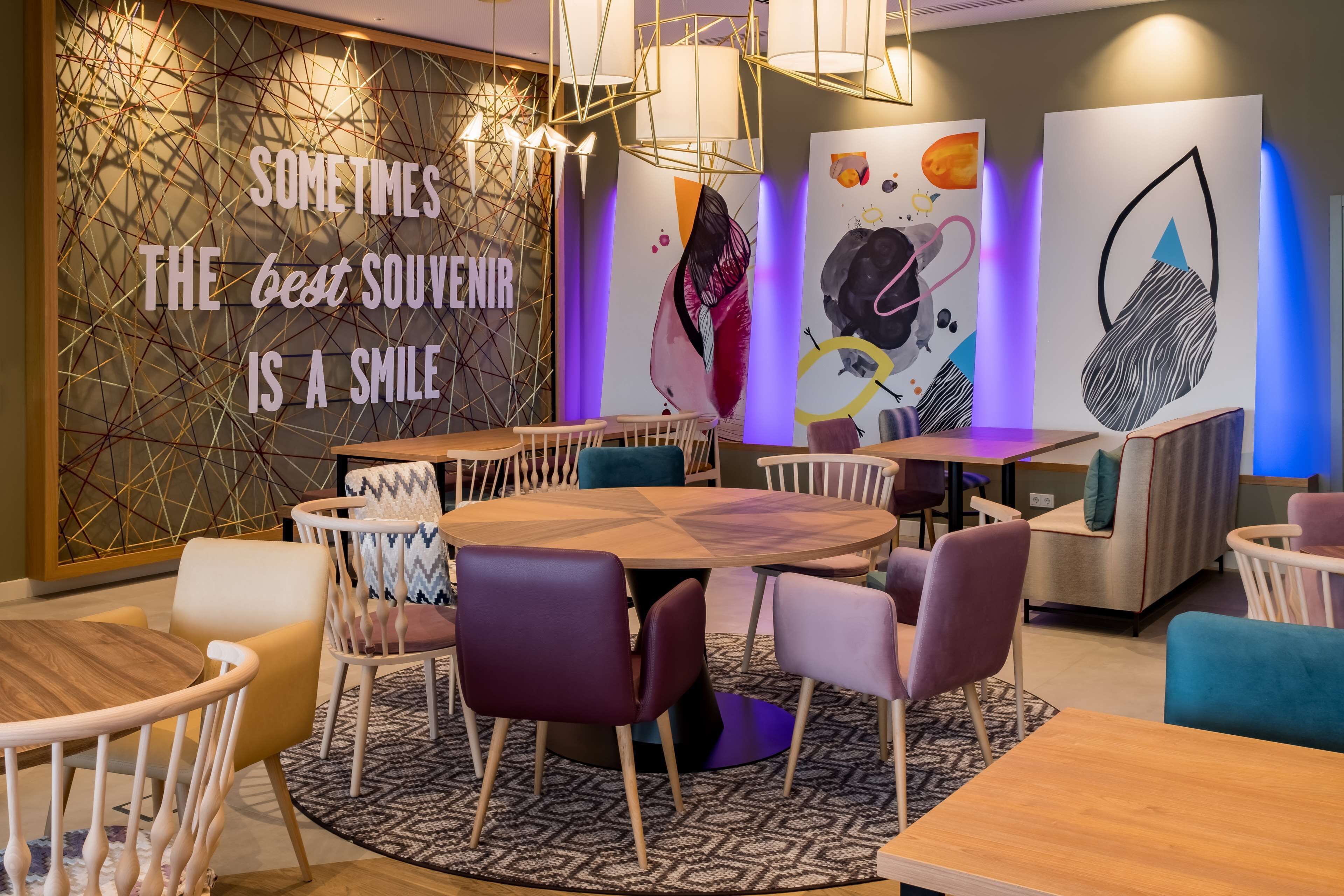 Uma área de estar moderna com uma mesa de madeira redonda rodeada por cadeiras coloridas, um painel decorativo com padrão geométrico e obras de arte abstratas.