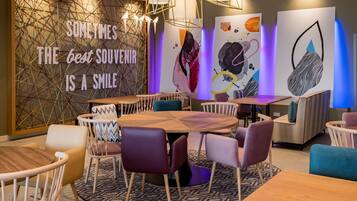 Uma área de estar moderna com uma mesa de madeira redonda rodeada por cadeiras coloridas, um painel decorativo com padrão geométrico e obras de arte abstratas.