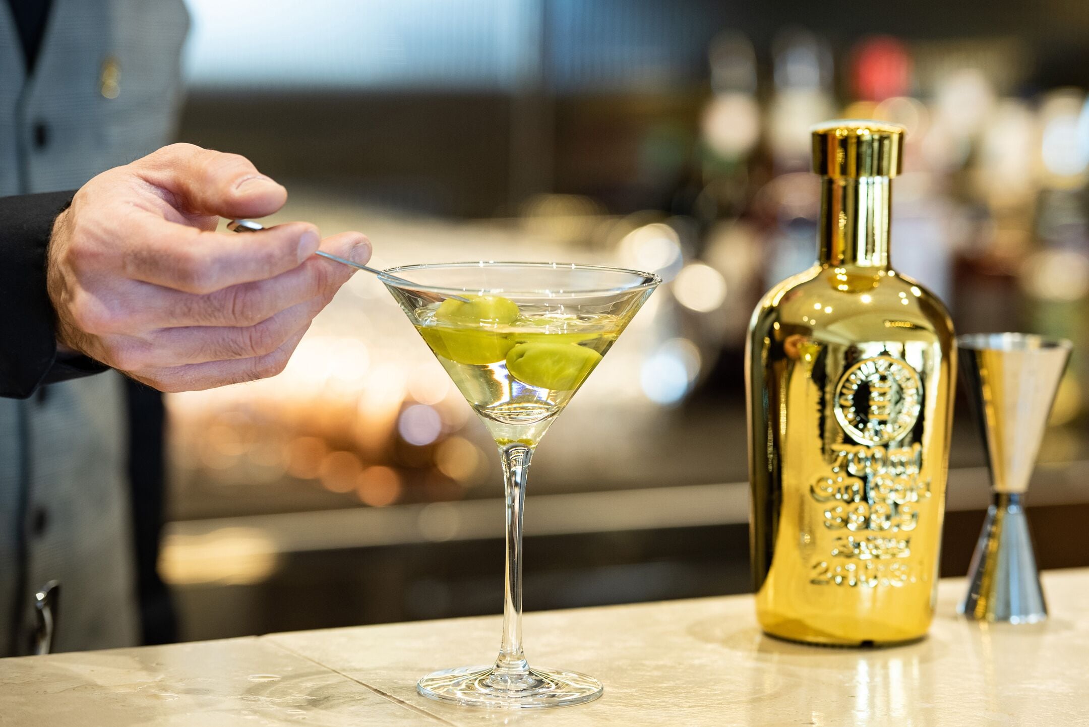 Um copo de martini com azeitonas em um balcão de bar ao lado de uma garrafa de bebida.