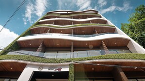 Edifício de vários andares com design moderno, com varandas ajardinadas e um logotipo em destaque na entrada.
