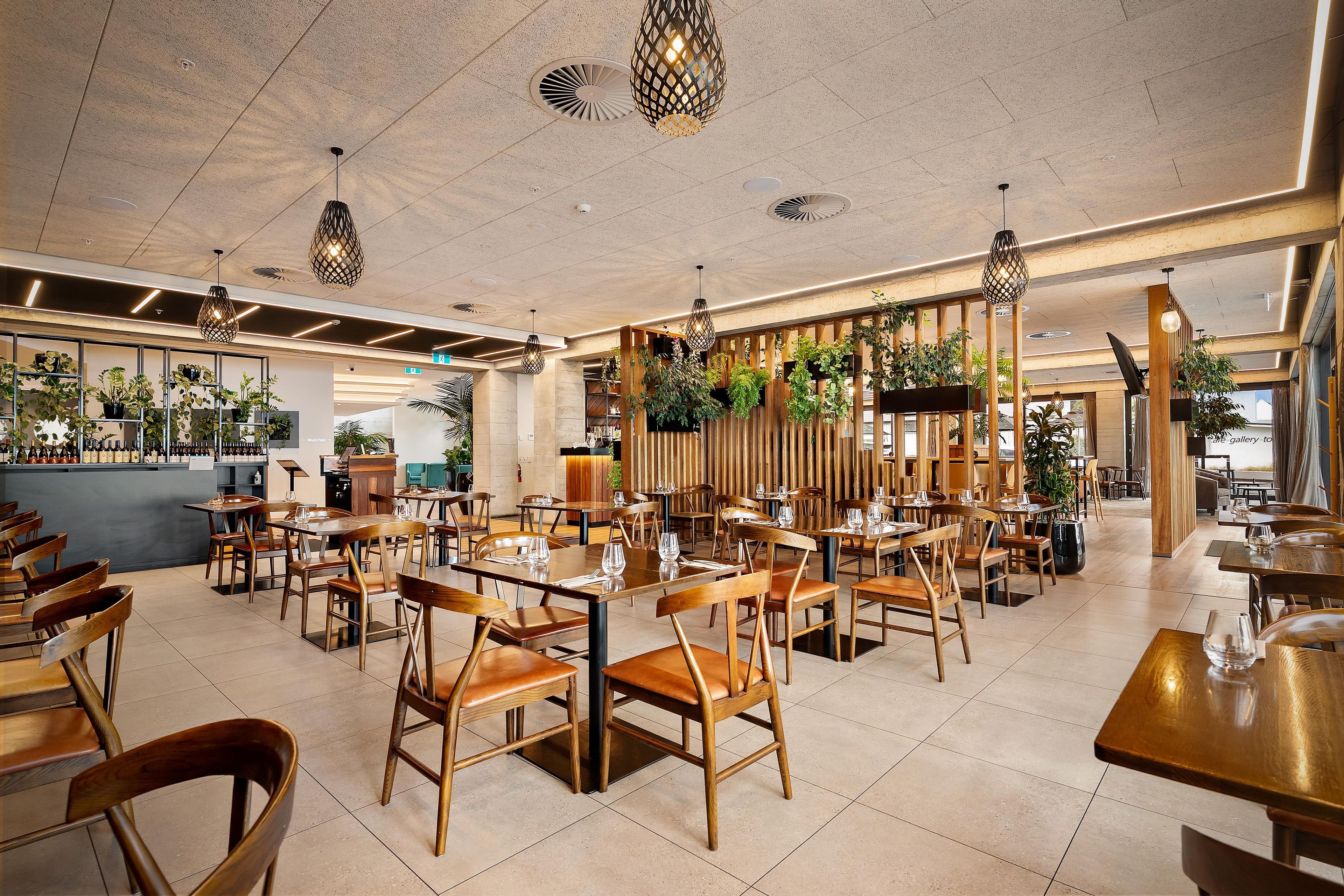 Um restaurante espaçoso com mesas e cadeiras de madeira, iluminação pendente e plantas em vasos.
