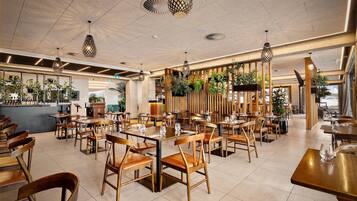 Um restaurante espaçoso com mesas e cadeiras de madeira, iluminação pendente e plantas em vasos.