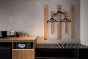 Quarto de hotel com escrivaninha de madeira, máquina de café, cofre e cabideiro fixado na parede.