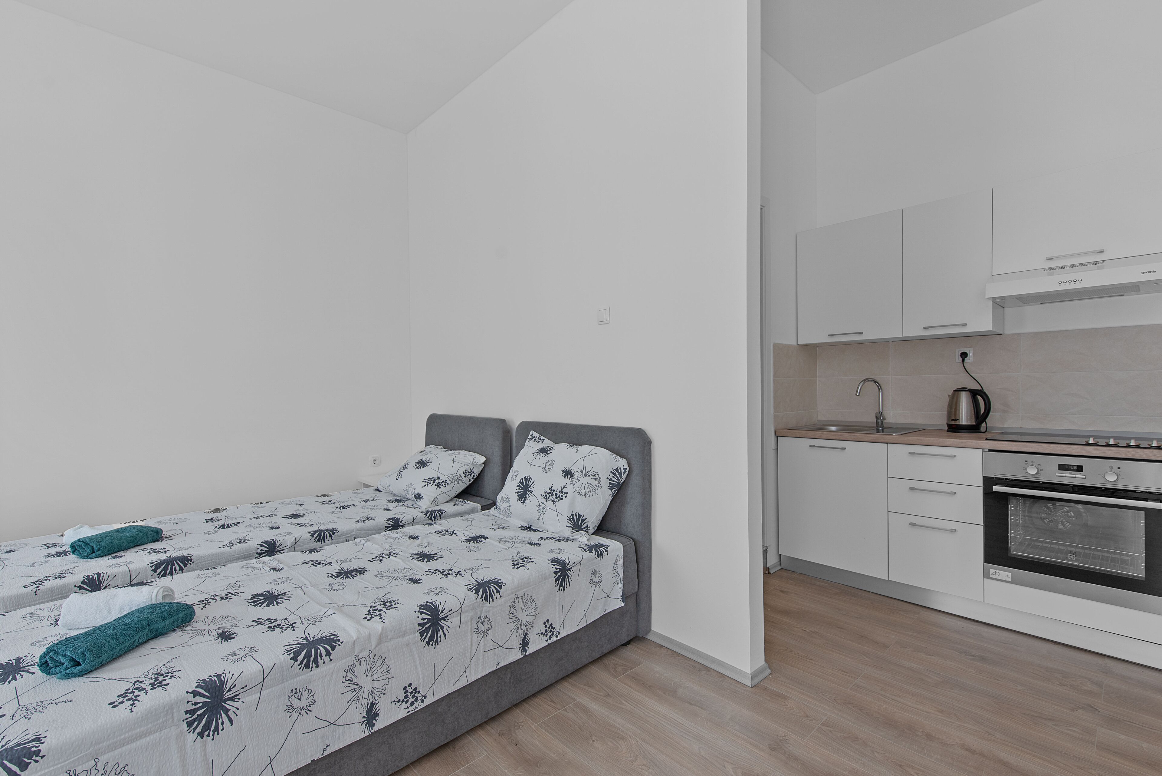 Quarto compacto com cama, duas almofadas e mesa de cabeceira. Um pequeno espaço de cozinha com fogão, forno e pia adjacente.