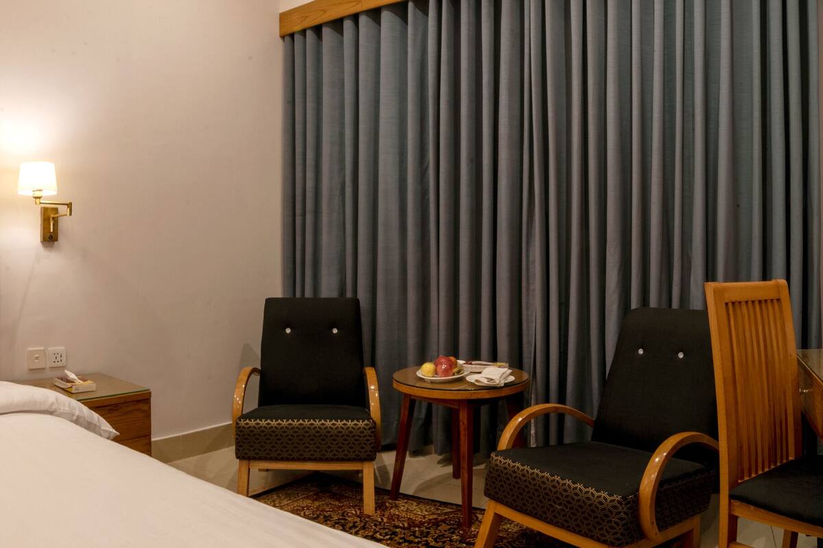 Quarto de hotel com uma cama grande, duas mesas de cabeceira, uma cadeira, uma pequena mesa com um prato e um copo, e um quadro na parede.