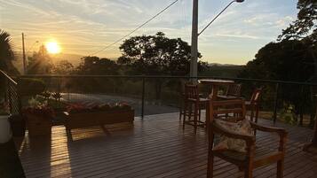 Um deck de madeira com mesa e cadeiras, com vista para uma estrada e árvores.