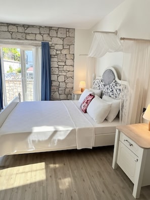 Quarto com parede de pedra, cama com roupa de cama branca, mesa de cabeceira com abajur e janela com cortinas.