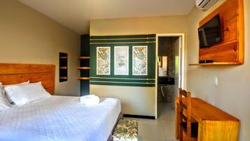 Quarto de hotel com uma cama, uma escrivaninha, uma cadeira, uma televisão e um banheiro visível por uma porta aberta.