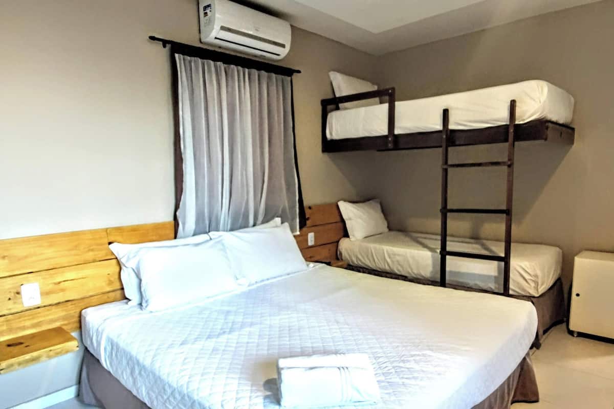 Quarto de hotel com uma cama grande, uma cama beliche, cabeceira de madeira, ar condicionado e uma pequena mesa de cabeceira.