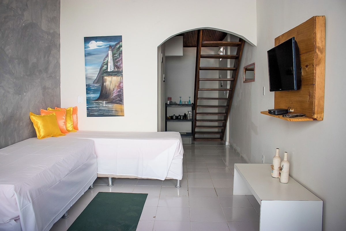 Quarto com cama, pintura, televisão e escrivaninha com garrafas.