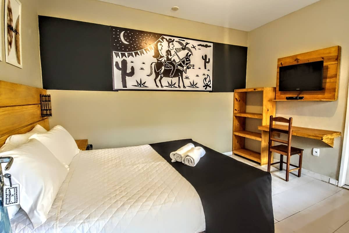 Quarto de hotel com uma cama, cabeceira de madeira, televisão presa a uma prateleira de madeira e um mural na parede.