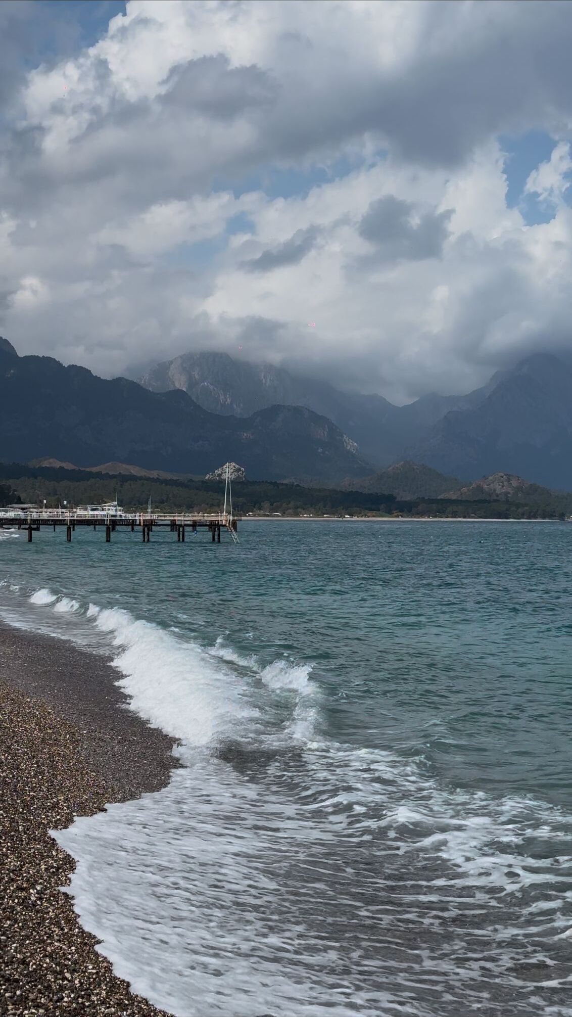 Uma praia com um cais que se estende por águas calmas, rodeada por montanhas e um céu nublado.