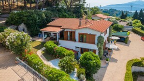 Uma casa de dois andares com telhado de telhas, cercada por vegetação exuberante e uma piscina.