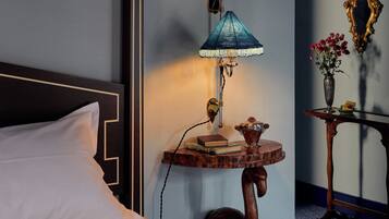Cama com travesseiro branco, luminária de parede com cúpula azul, mesinha com vaso de flores e espelho com moldura dourada.