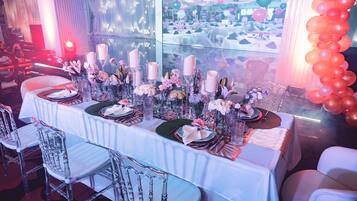 Salão de festas com mesas montadas para um evento formal, com cadeiras transparentes, toalhas brancas e arranjos florais.