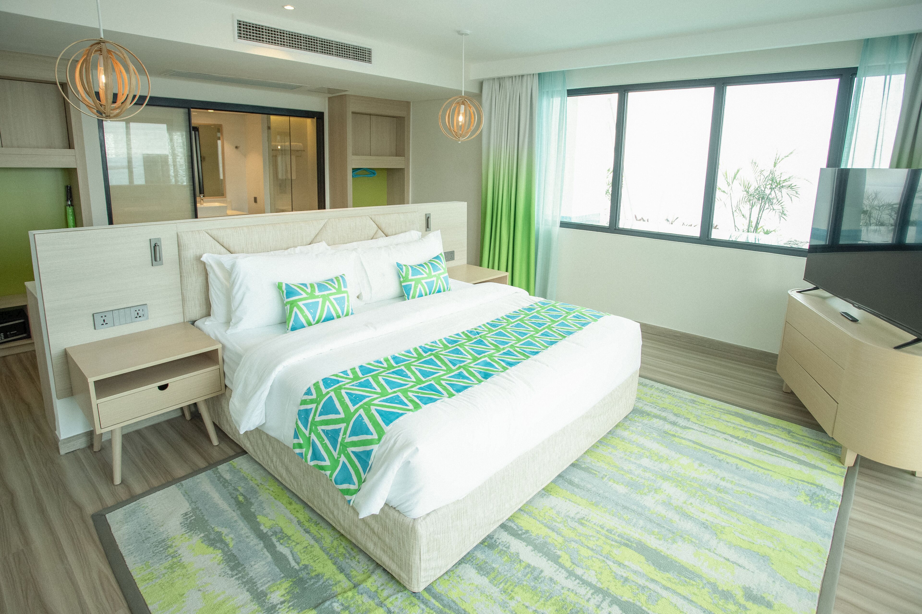 Quarto de hotel moderno com uma cama grande, televisão, mesa de cabeceira e janela com vista.