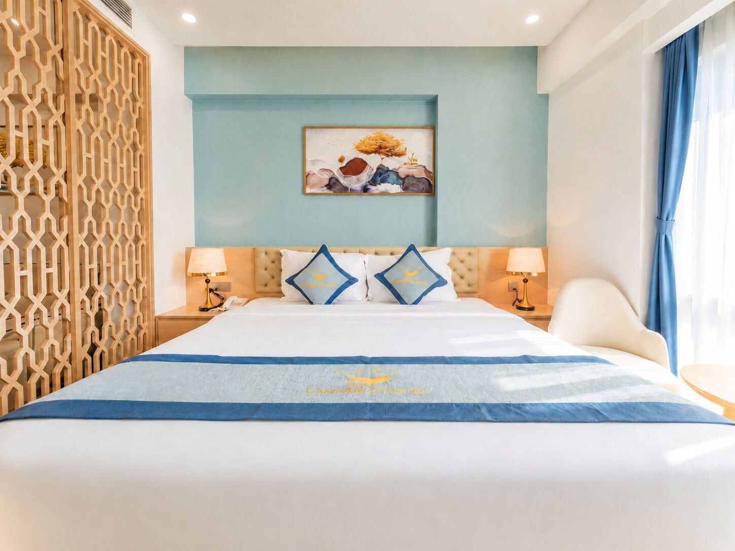 Quarto de hotel com uma cama, duas mesas de cabeceira, uma cadeira, um painel decorativo na parede e uma obra de arte emoldurada acima da cama.