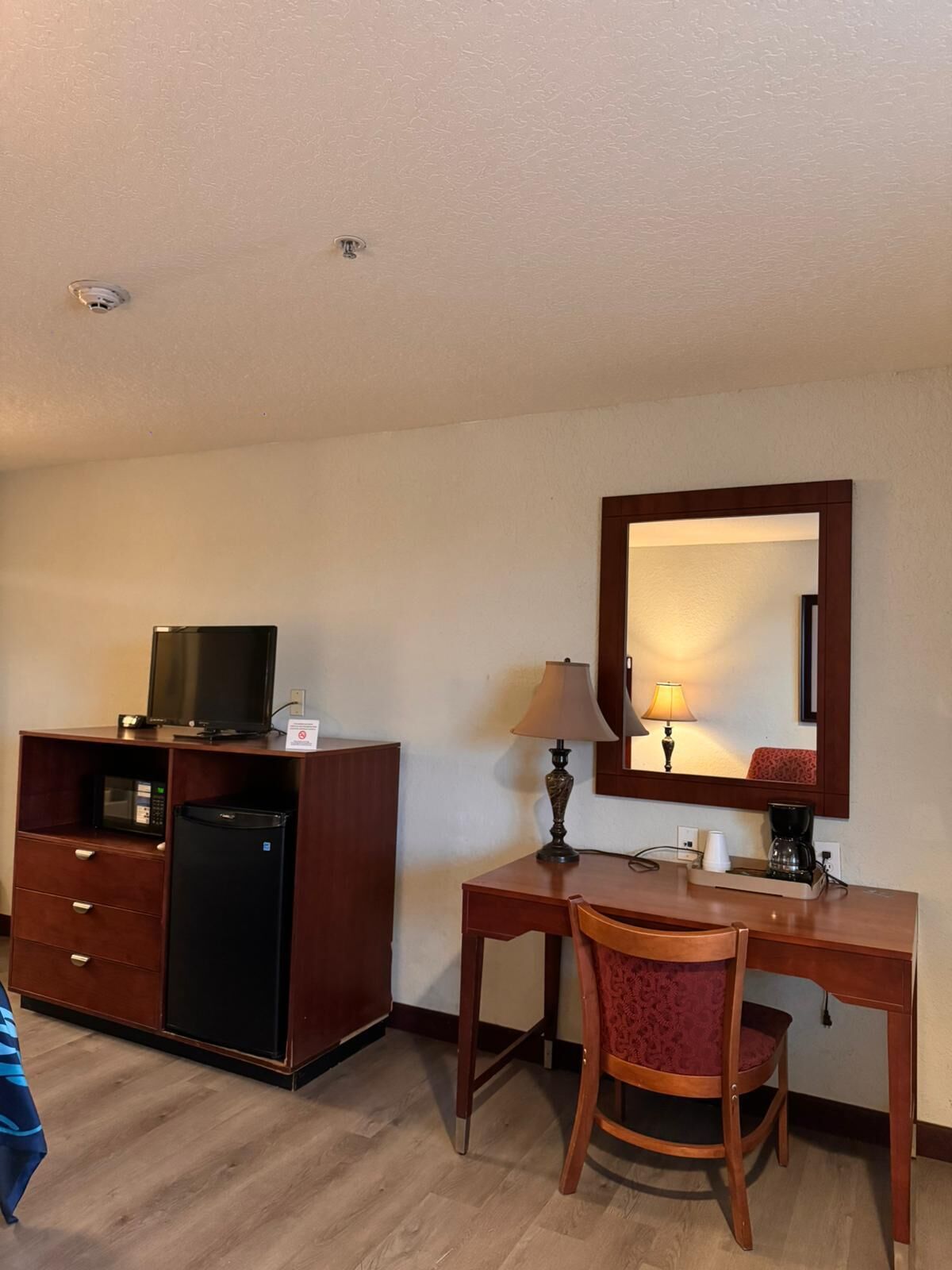 Quarto de hotel com uma escrivaninha de madeira, uma cadeira, televisão, frigorífico, micro-ondas, candeeiro e espelho.