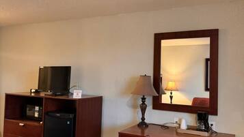 Quarto de hotel com uma escrivaninha de madeira, uma cadeira, televisão, frigorífico, micro-ondas, candeeiro e espelho.