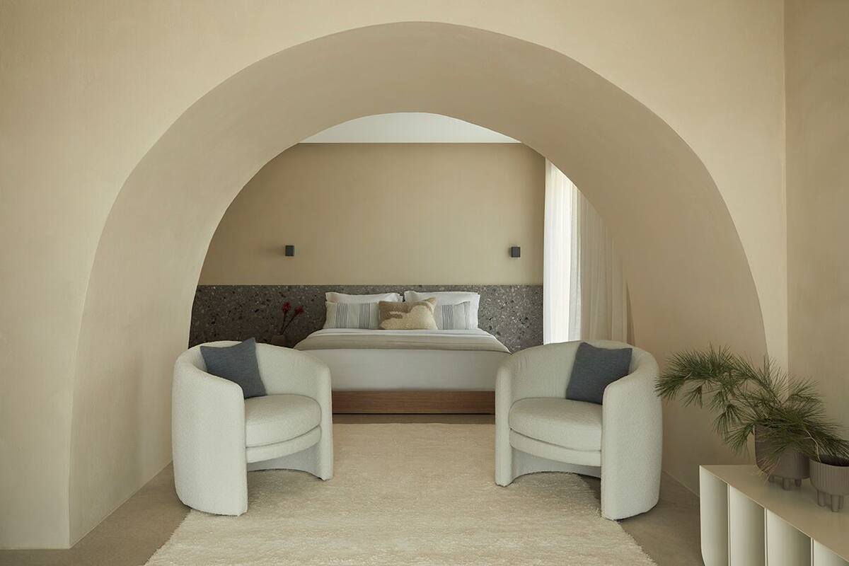 Quarto com uma grande porta em arco, cama com roupa de cama branca, duas poltronas brancas e um pequeno vaso com planta.