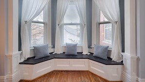 Janela baía com cortinas brancas, duas almofadas e piso de madeira.