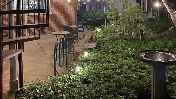 Uma área de pátio externa com luzes de fada, um edifício de tijolos e vegetação.