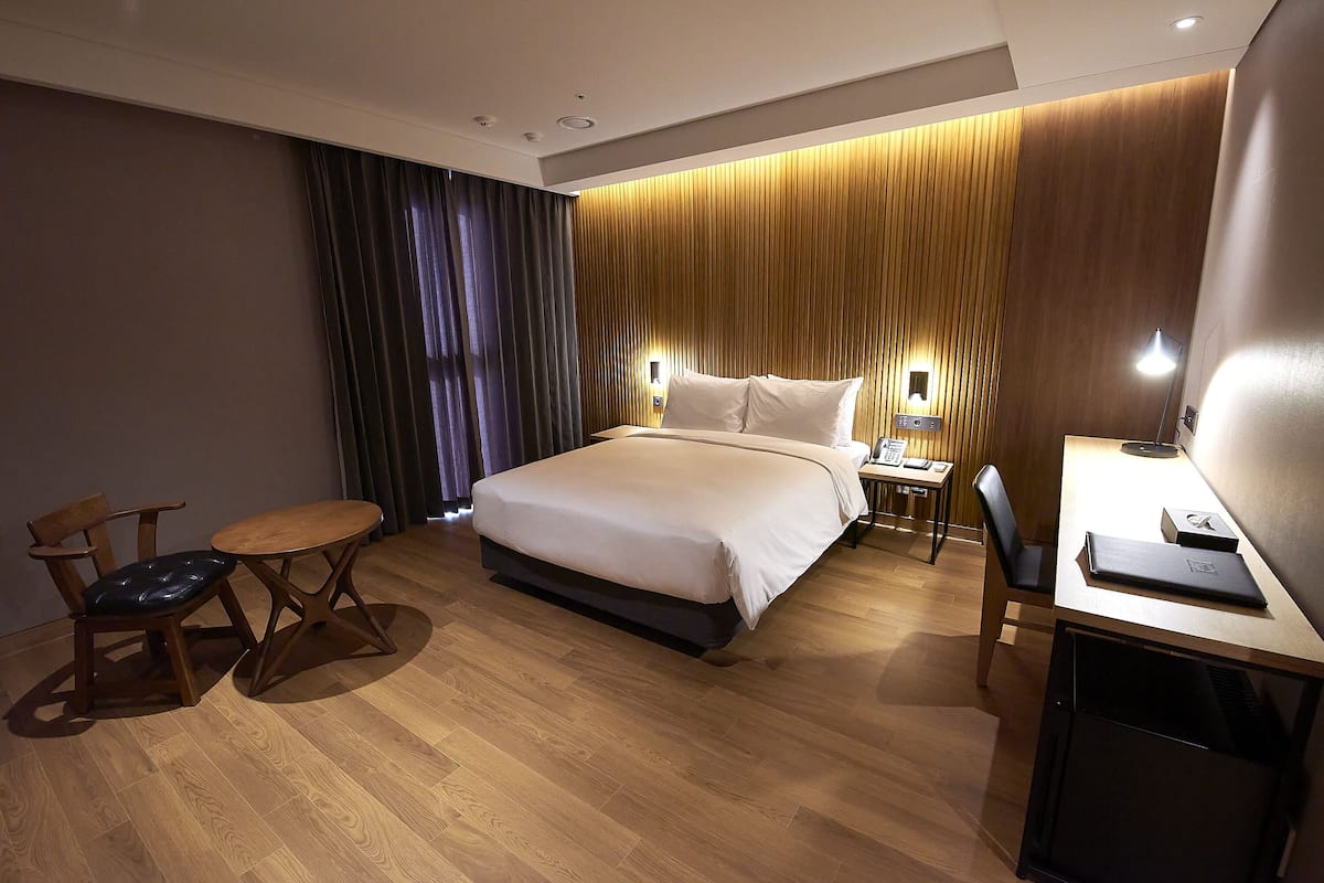 Quarto de hotel com cama grande, escrivaninha, cadeira e mesa pequena.