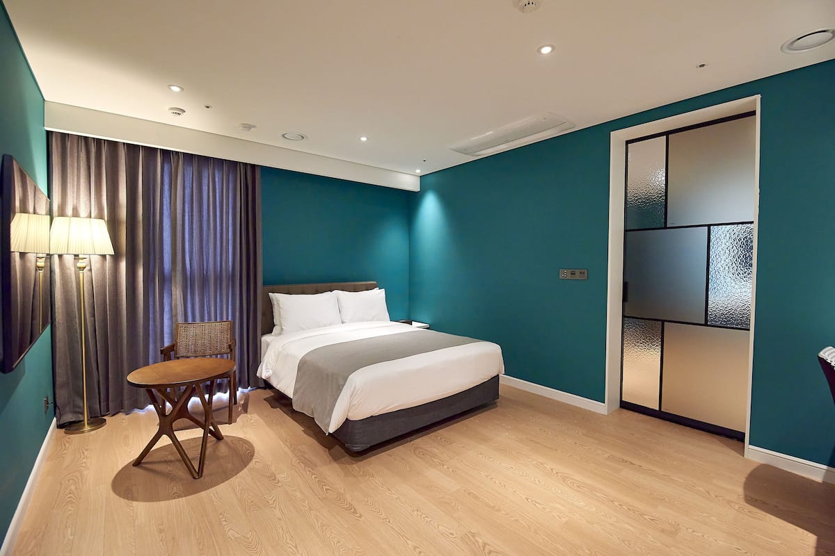 Quarto de hotel moderno com cama, uma cadeira, abajur e uma janela grande com cortinas.