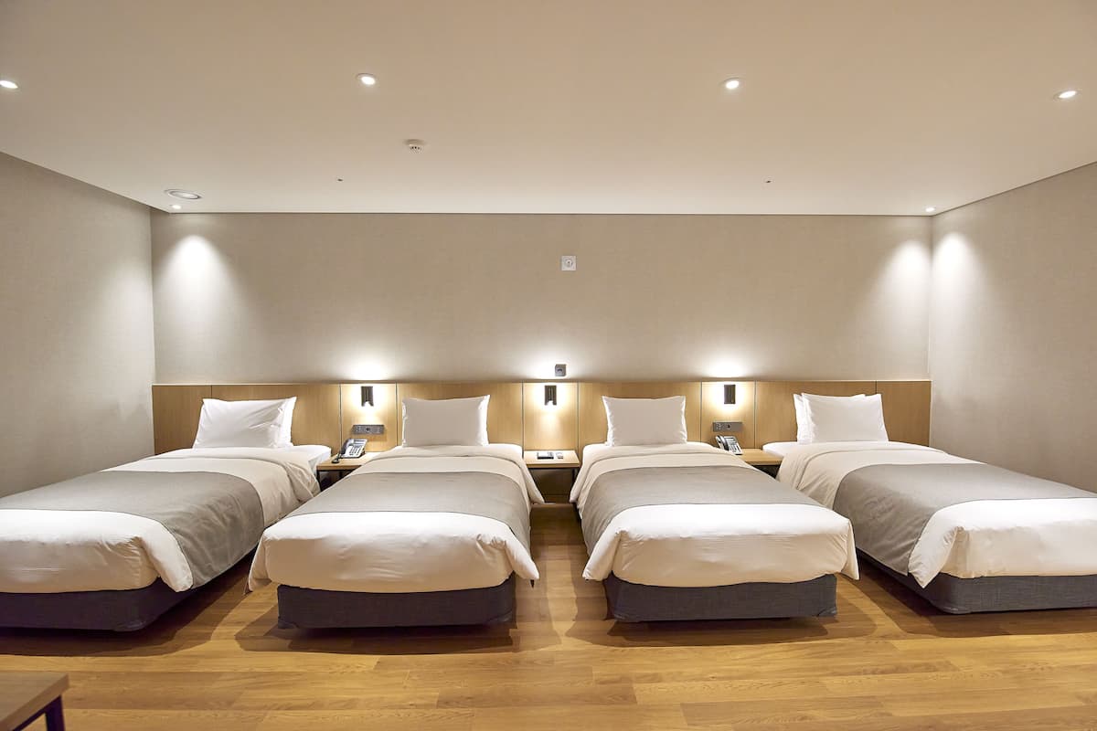 Quarto de hotel com quatro camas de solteiro dispostas em fila, cabeceiras de madeira e iluminação embutida no teto.