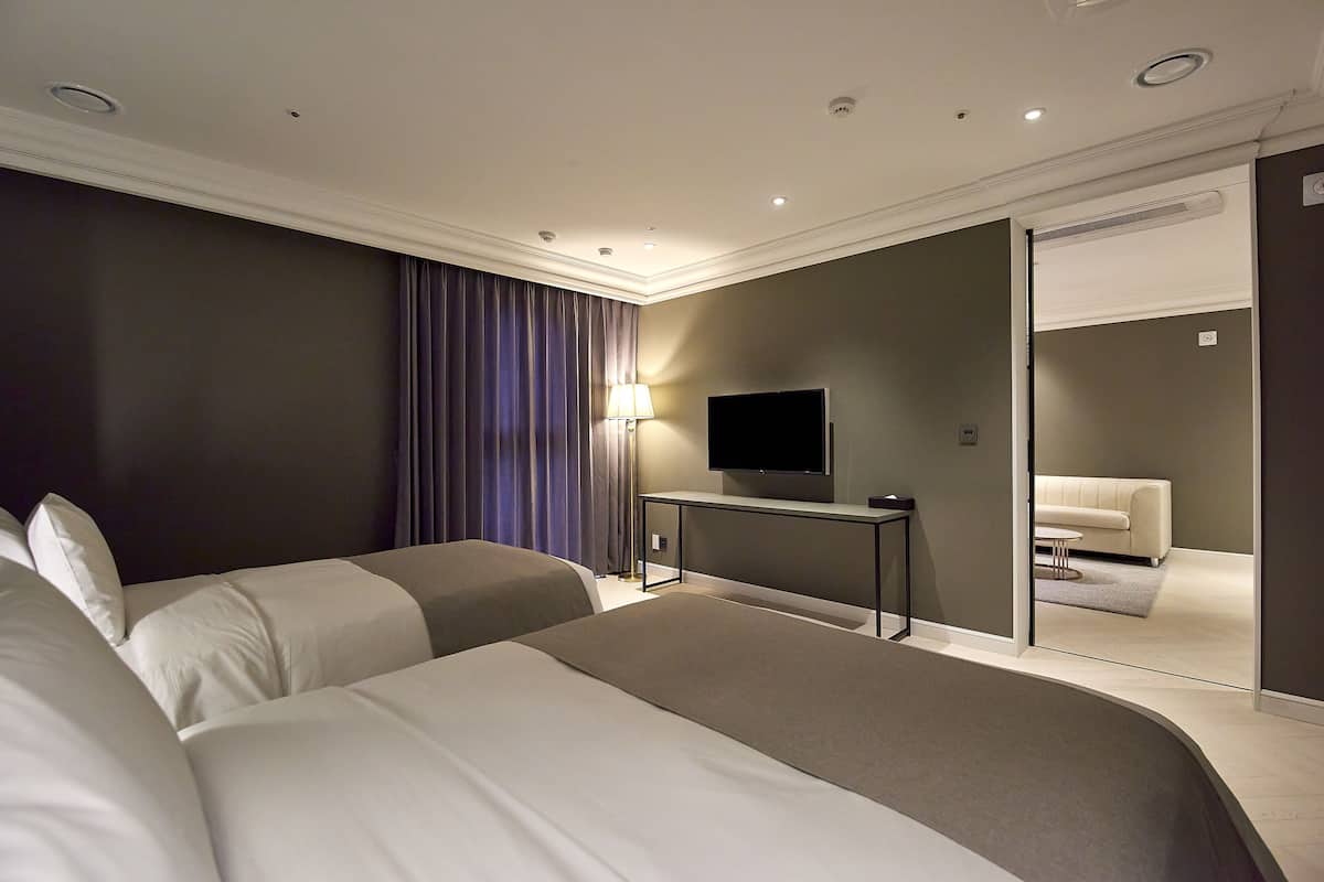 Quarto de hotel moderno com duas camas, televisão, escrivaninha e vista para uma sala de estar.