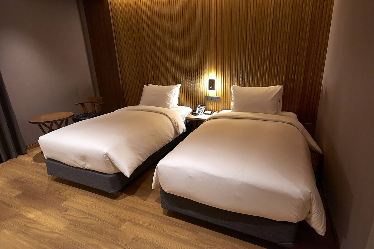 Uma cama de casal com cabeceira de madeira, uma mesa de cabeceira com abajur e um banquinho de madeira.