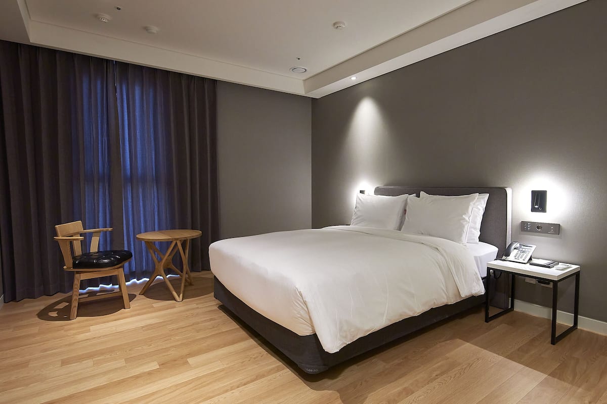 Quarto de hotel moderno com uma cama grande, uma mesinha com telefone e uma cadeira.