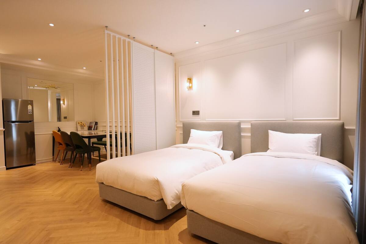 Quarto de hotel com duas camas, área de refeições com cadeiras, geladeira e luminária fixada na parede.