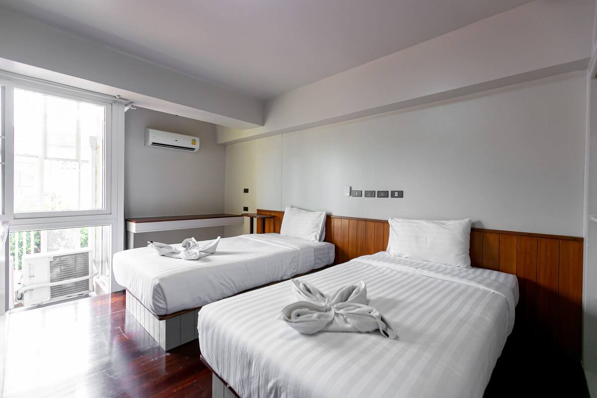 Quarto de hotel com duas camas, roupa de cama branca, cabeceiras de madeira, janela com unidade de ar condicionado e vista para a varanda.