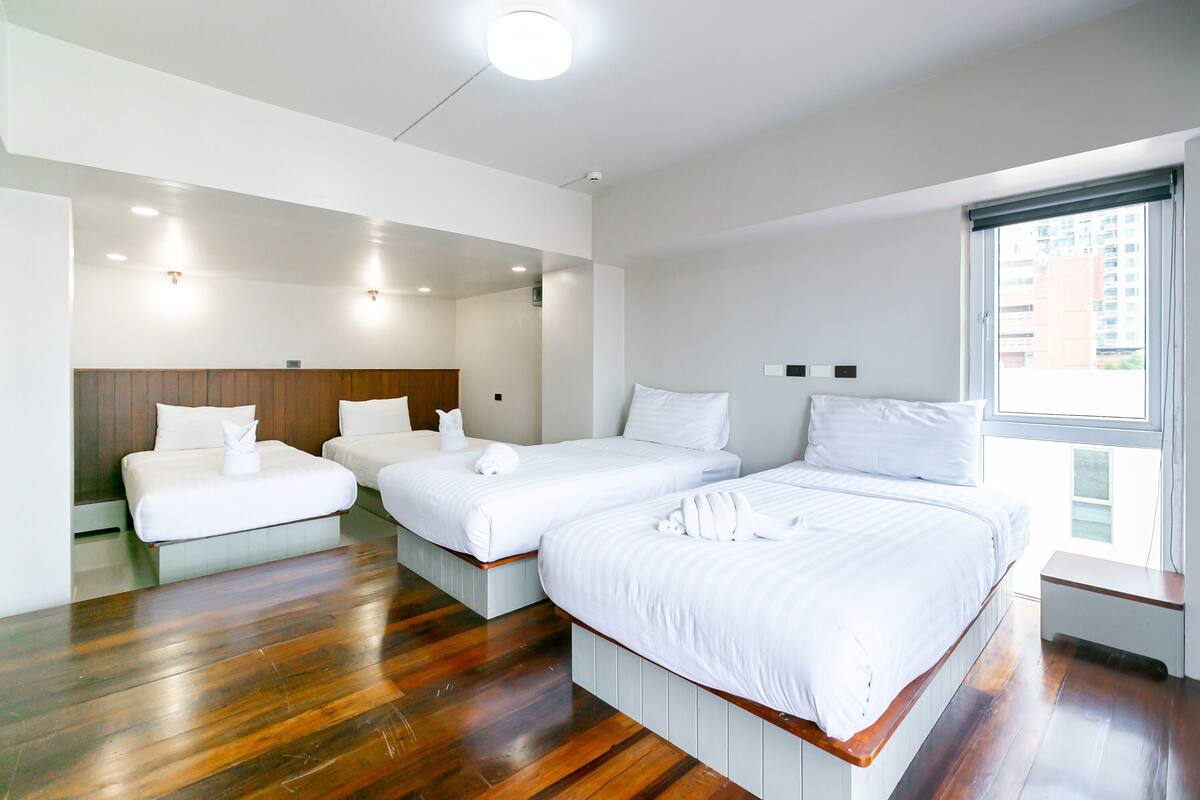 Quarto de hotel com três camas, piso de madeira e uma janela grande com vista para a cidade.